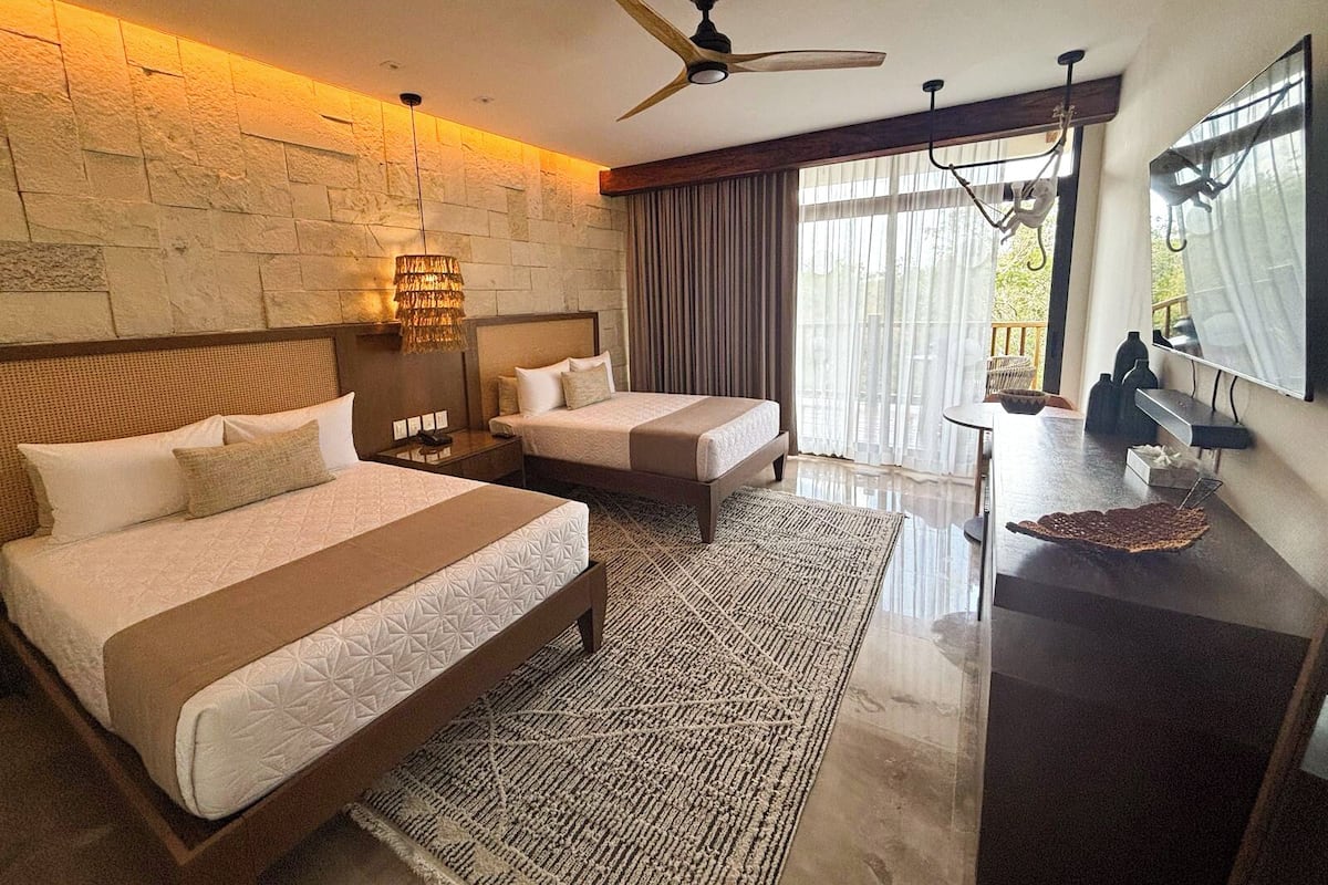 Quarto de hotel com uma cama grande, uma cama menor, uma escrivaninha e uma varanda com cortinas.