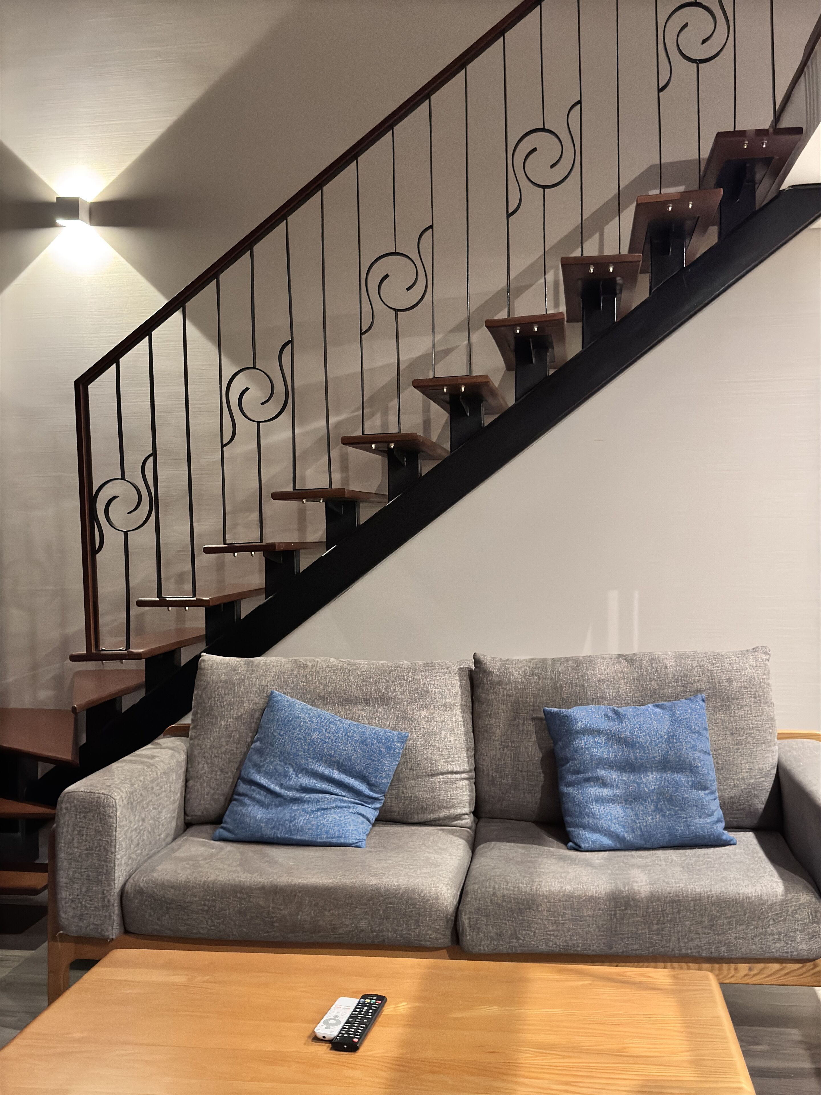 Uma sala de estar moderna com um sofá cinza, duas almofadas azuis, uma mesa de centro de madeira e uma escada preta com guarda-corpo decorativo.