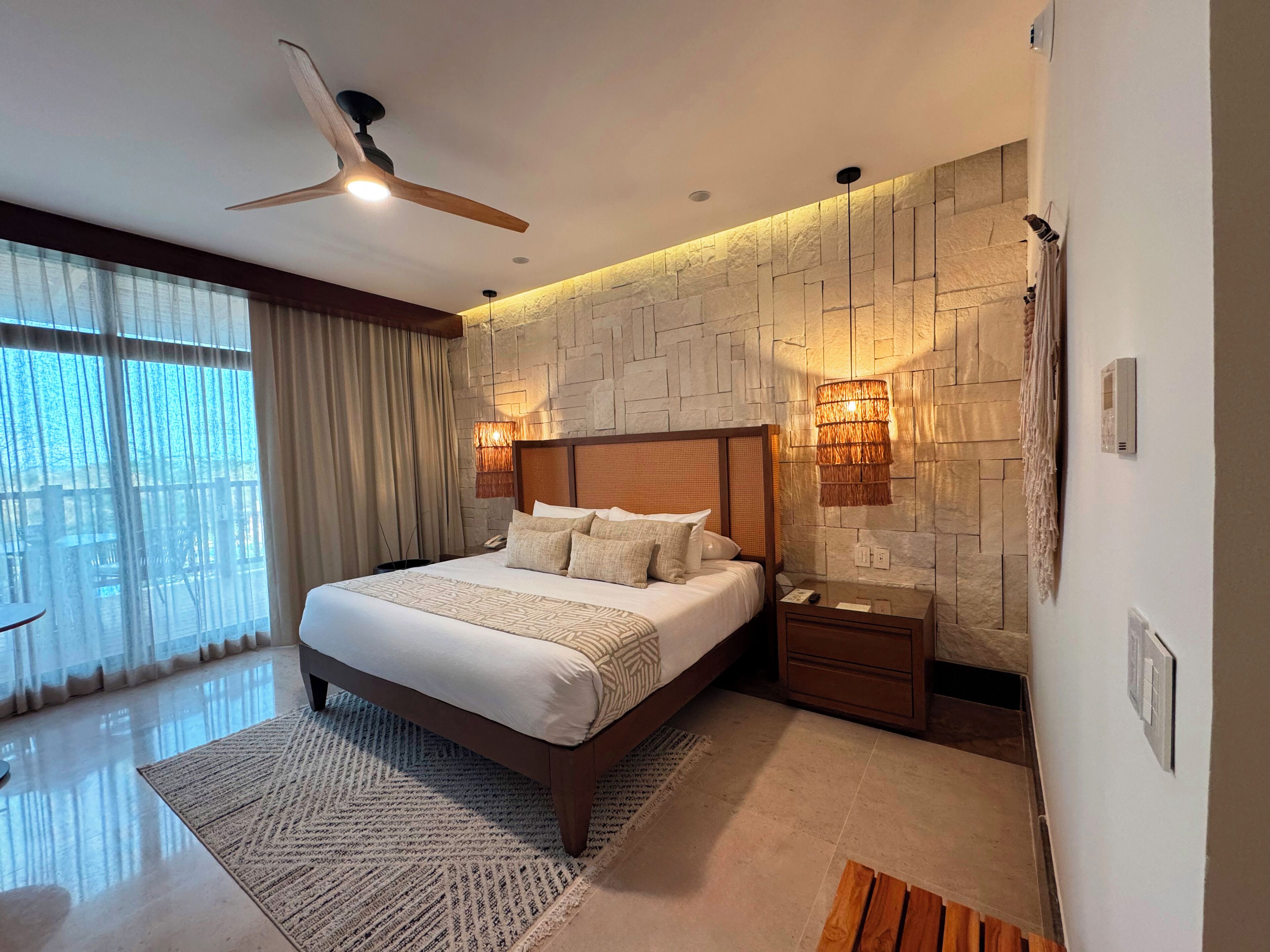 Quarto de hotel moderno com uma cama grande, um ventilador de teto, parede com padrão de pedra e vista para o exterior.