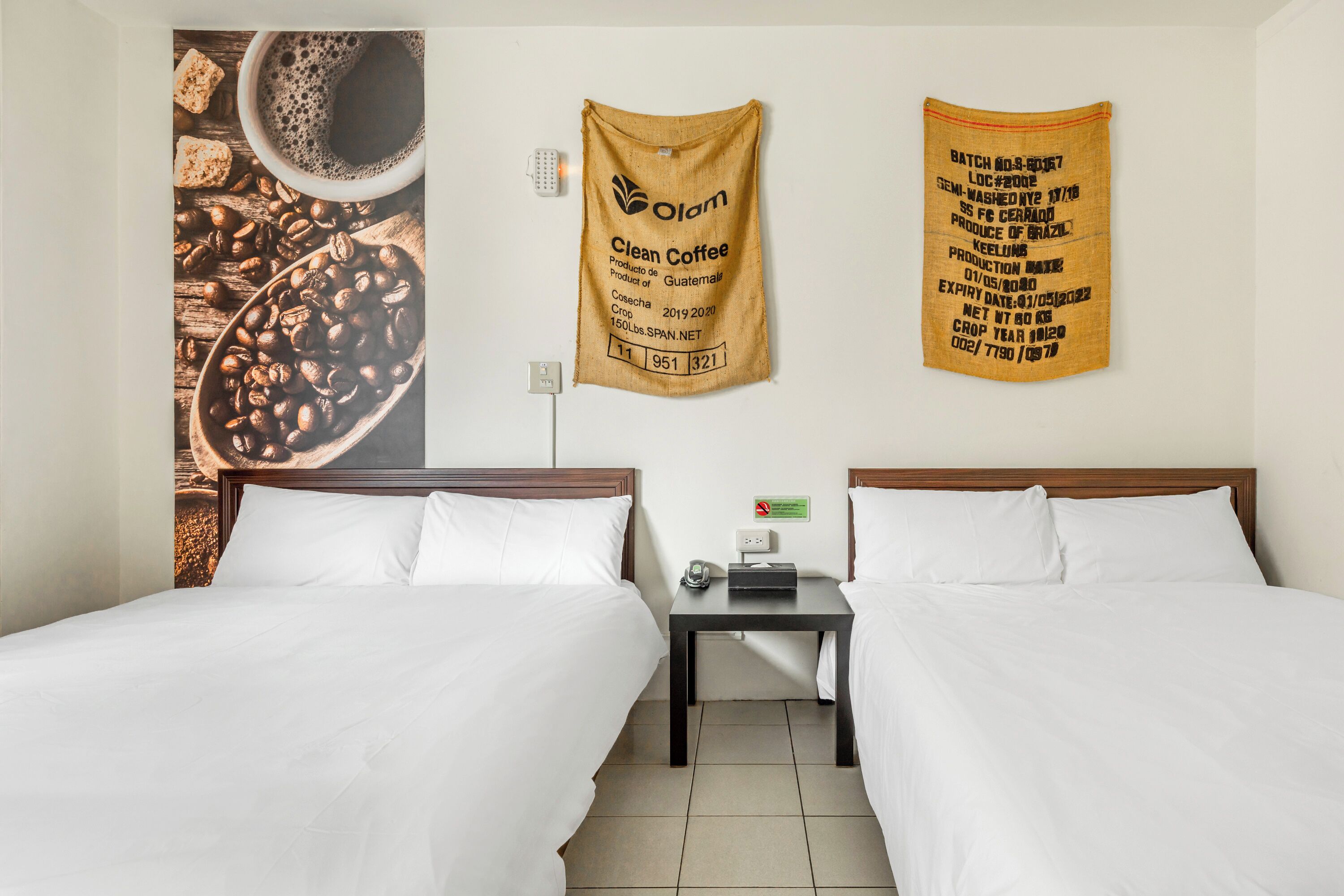 Quarto de hotel com duas camas, uma mesa de cabeceira e parede decorada com cartazes sobre café.