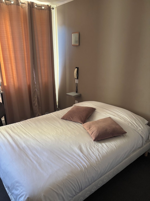 Quarto de hotel com uma cama, duas almofadas, mesa de cabeceira com telefone e uma janela com cortinas.