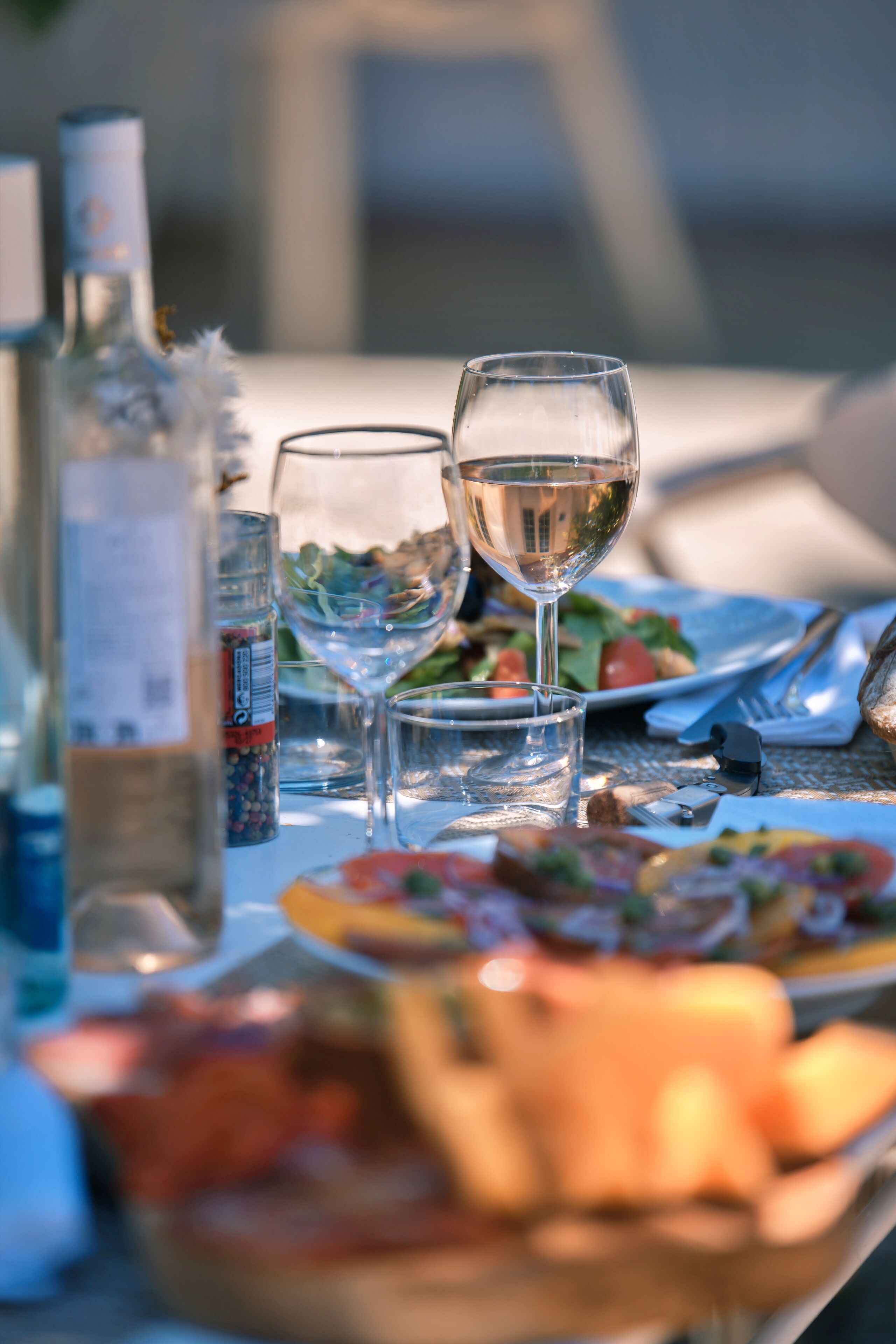 Uma mesa posta com uma garrafa de vinho branco, duas taças de vinho, uma salada e um prato de pão.