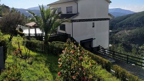 Uma casa de dois andares com fachada branca e telhado de cerâmica, rodeada por vegetação exuberante e uma paisagem montanhosa.