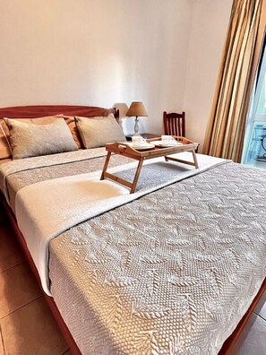 Uma cama bem arrumada com um cobertor estampado, uma bandeja de madeira com xícaras e pires, e um abajur em uma mesa de cabeceira.
