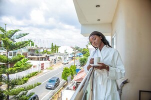 Alguém numa varanda segurando uma xícara, com vista para uma rua com carros e edifícios.