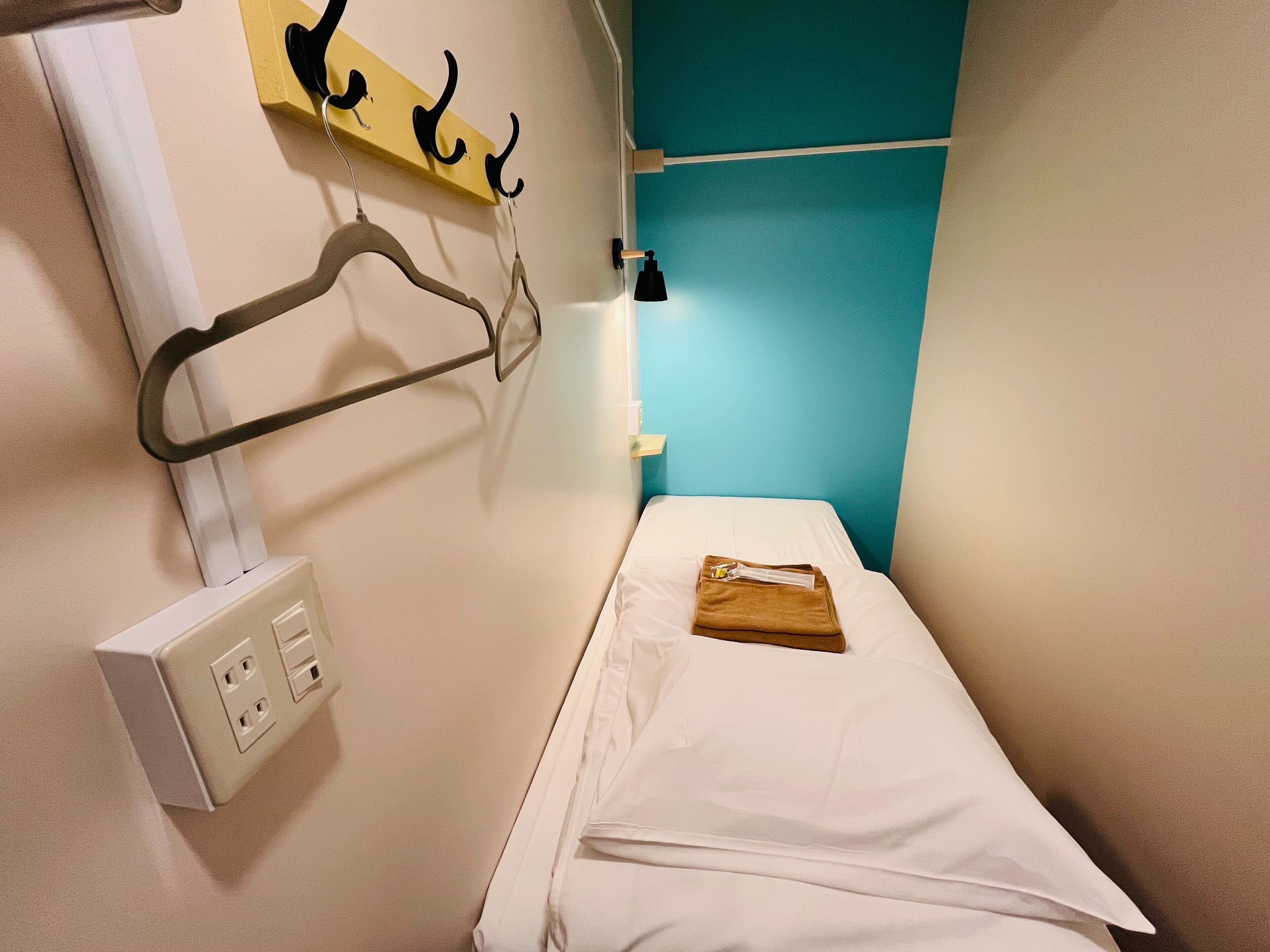 Quarto pequeno, com uma cama de solteiro, colchão branco, mesa de cabeceira de madeira, luminária na parede e um gancho para casacos.