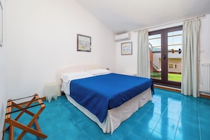 Quarto com cama, uma cadeira de madeira, janela com cortinas e vista para um jardim.