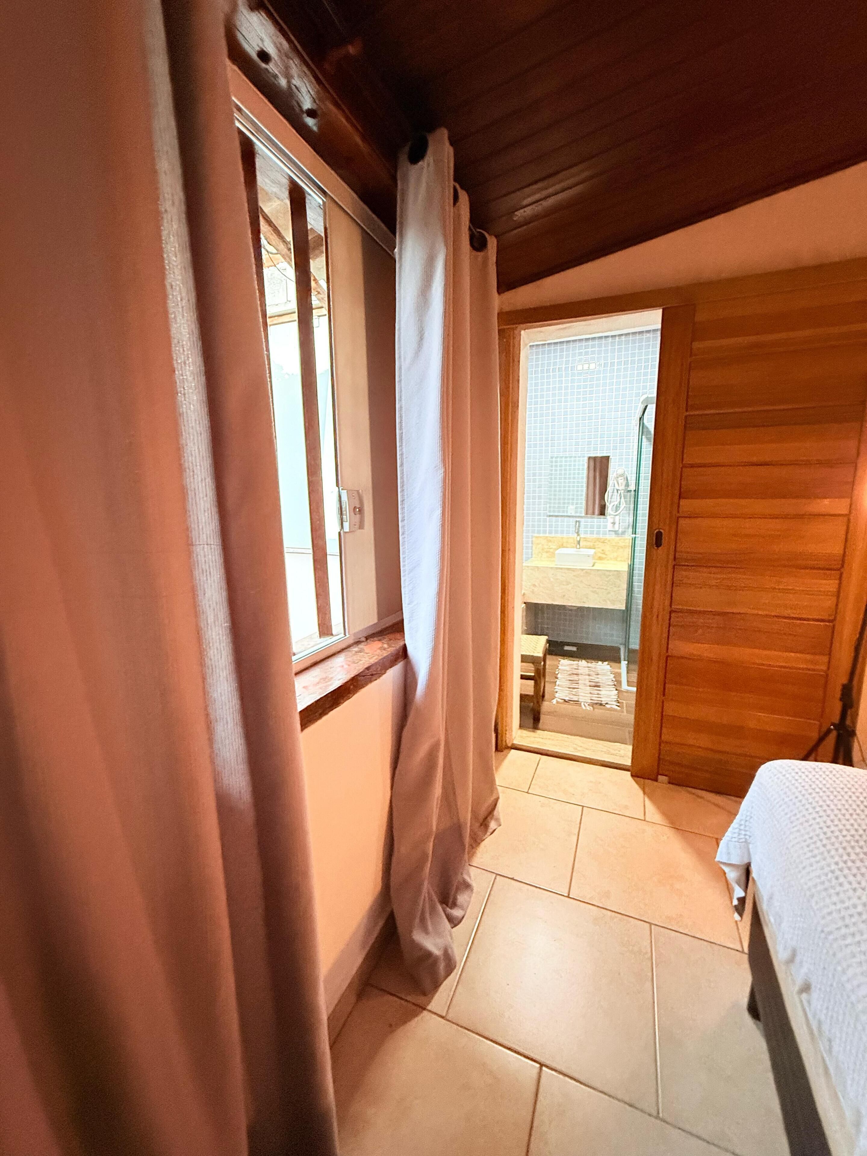 Quarto com porta de madeira, janela com cortinas e piso ladrilhado.