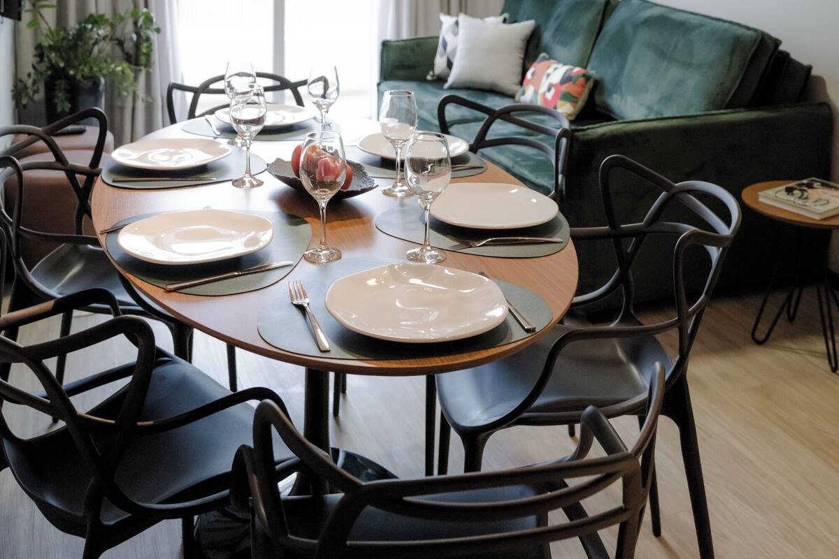 Uma área de refeições com uma mesa redonda posta para quatro pessoas, rodeada por cadeiras pretas. Há um sofá verde com uma almofada e uma pequena mesa lateral com uma luminária. Uma grande janela com cortinas é visível ao fundo.