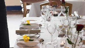 Mesa de jantar posta com copos, pratos e talheres, com uma pessoa organizando a disposição.