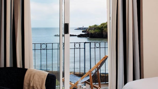 Quarto com uma grande janela com vista para a costa, uma cama, uma cadeira e uma pequena mesa com uma garrafa.