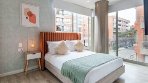 Quarto moderno com uma cama grande, mesa de cabeceira com luminária e vista da cidade através de grandes janelas.