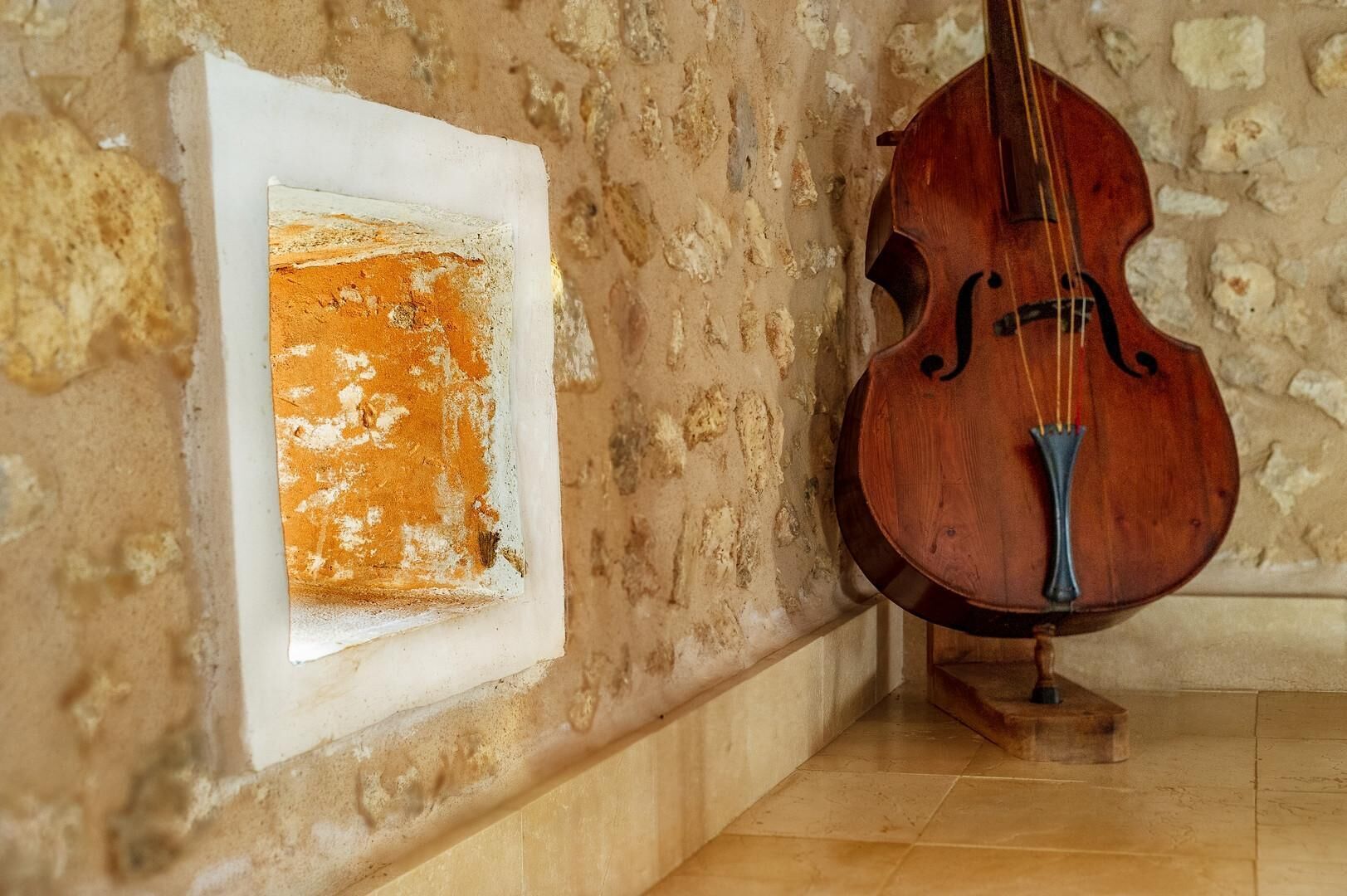 Um violoncelo encostado numa parede de pedra texturizada, com uma obra de arte emoldurada com um design abstrato em laranja e branco.