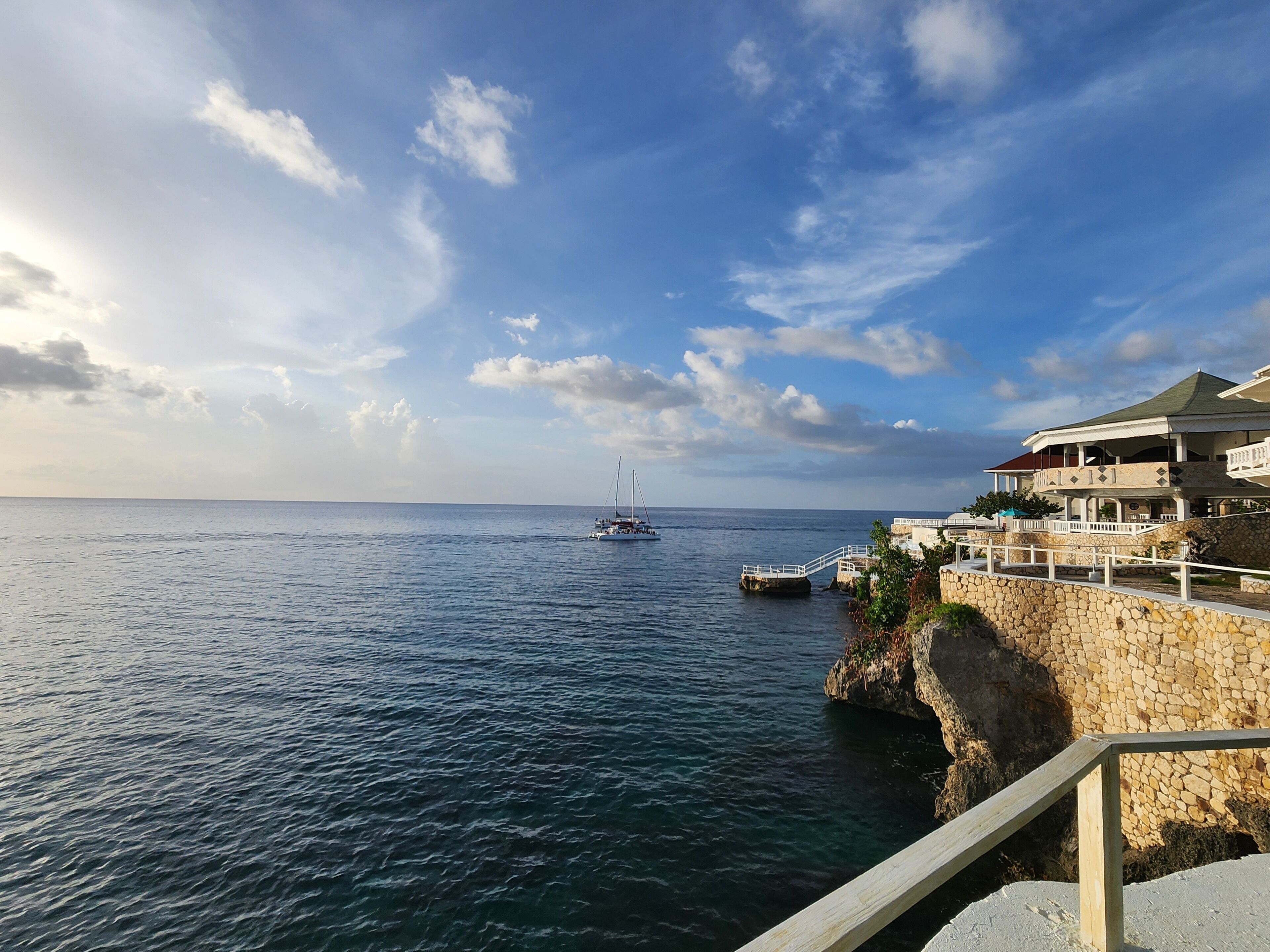 Uma cena costeira com um cais de pedra que leva a um edifício com varanda com vista para o mar.