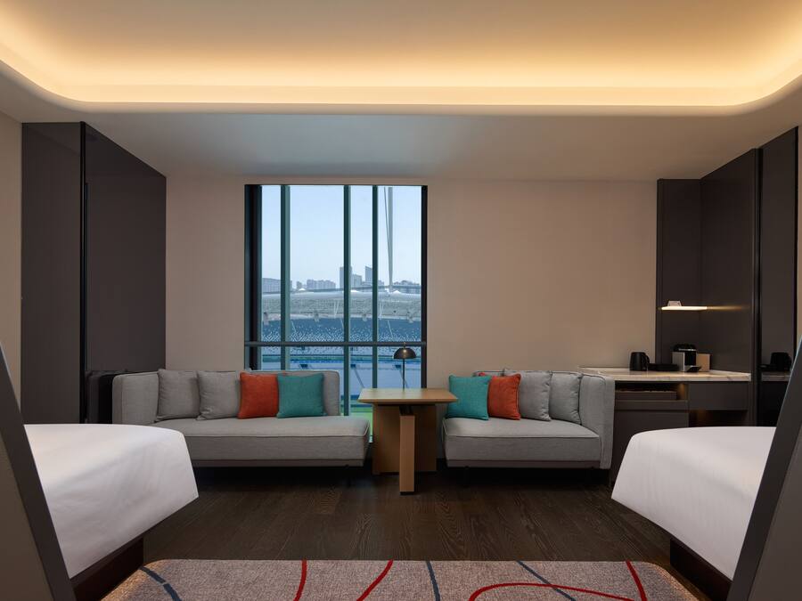 Quarto de hotel com duas camas, um sofá, uma mesinha e vista para a paisagem urbana.