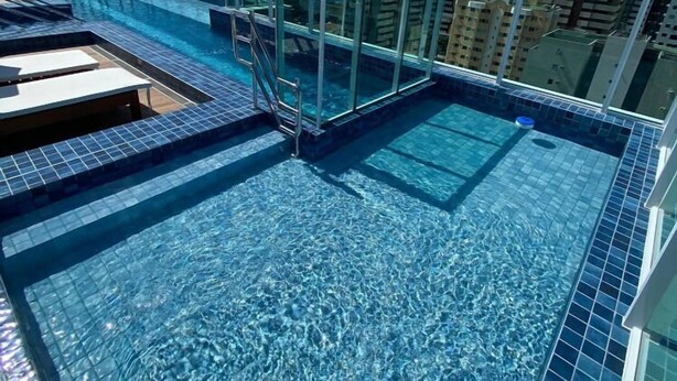 Piscina no terraço com água azul cristalina, guarda-corpos de vidro e vista para a paisagem urbana ao fundo.