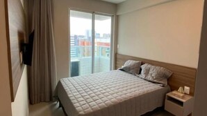 Quarto de hotel com uma cama, mesa de cabeceira, ar-condicionado fixado na parede e vista para a cidade através de uma grande janela.