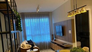 Uma sala de estar moderna com sofá, mesa de centro e área de jantar. Há uma TV na parede e uma escada com uma planta ao lado.