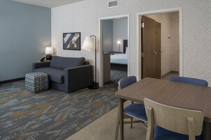 Quarto de hotel com sofá, mesa pequena, luminária, tapete estampado e cama com cabeceira.