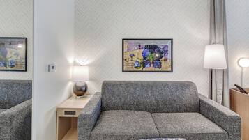 Quarto de hotel moderno com um sofá cinza, uma pequena mesa lateral de madeira, uma luminária e uma obra de arte emoldurada na parede.
