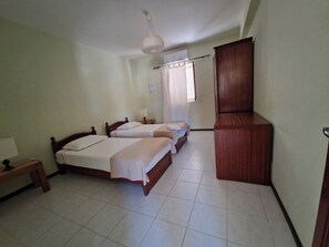 Quarto com duas camas de madeira, uma pequena mesa com luminária, um armário de madeira e um ventilador de teto.