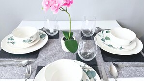 Mesa posta com pratos e tigelas brancos com padrões de folhas verdes, copos de vidro transparentes e uma orquídea em vaso.