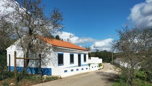 Um edifício branco com detalhes em azul e telhado vermelho, rodeado por vegetação e um caminho de terra.