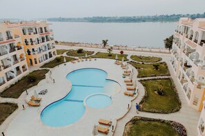 Piscina com design exclusivo, rodeada por espreguiçadeiras e guarda-sóis, com vista para um lago e edifícios ao fundo.