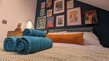 Quarto com cama, toalhas azuis dobradas, tapete estampado e parede decorada com pôsteres de viagens.