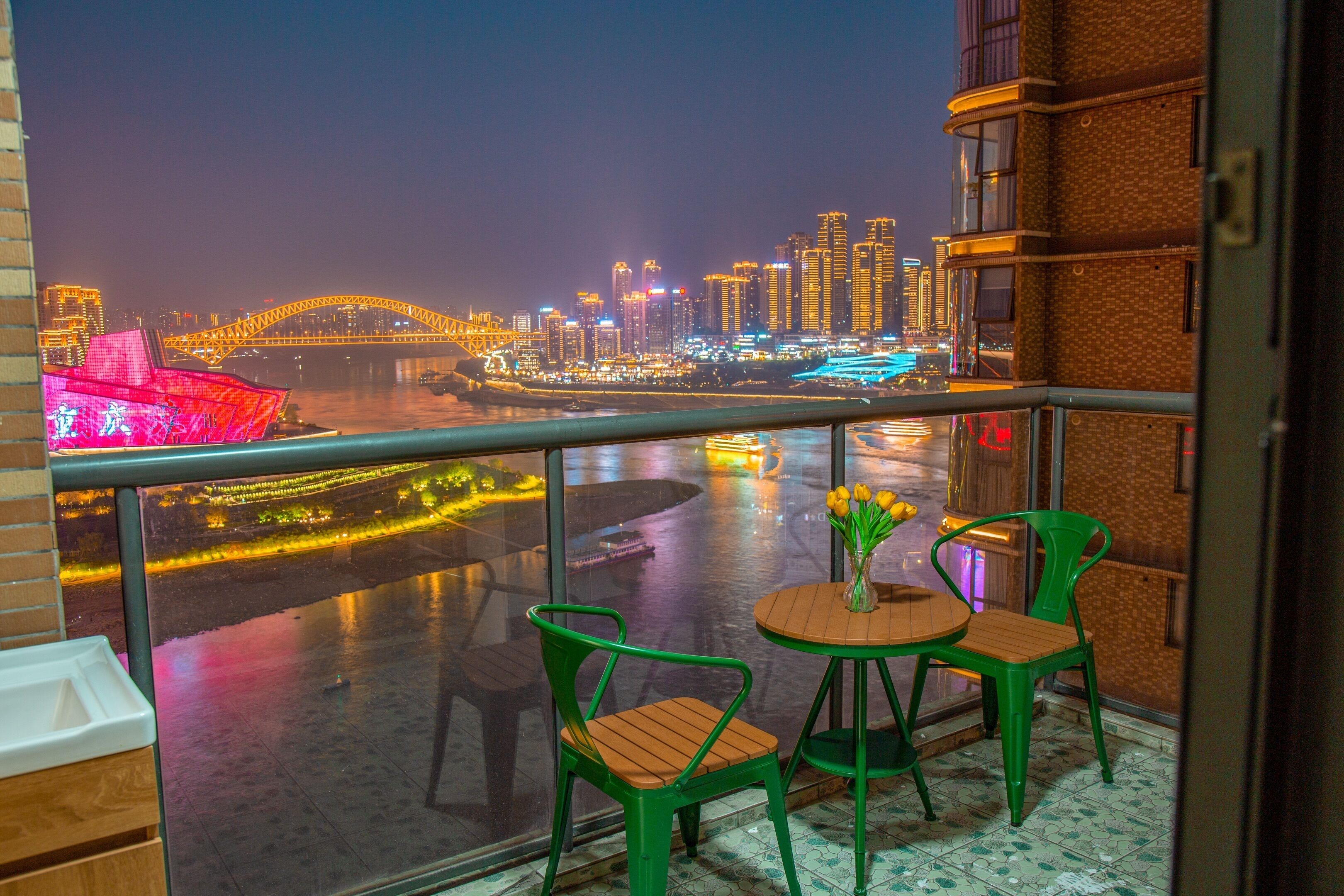 Varanda com duas cadeiras verdes e uma mesa pequena, com vista para uma paisagem urbana com edifícios iluminados e uma ponte.