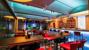Uma área de bar com mesas e cadeiras vermelhas, uma tela de televisão e prateleiras com garrafas.