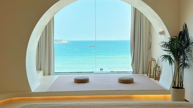 Quarto com uma grande janela arqueada com vista para o mar, uma pequena mesa com almofada e um vaso com planta.