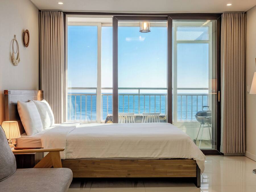 Quarto de hotel com cama, sofá, escrivaninha, luminária, churrasqueira e vista para o mar.