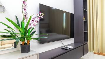 Uma sala de estar moderna com uma grande TV de tela plana fixada em uma parede escura, uma planta em vaso com folhas verdes e flores roxas, e um sofá bege.