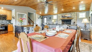 Uma sala de jantar com teto de madeira, lareira de pedra e cozinha ao fundo.
