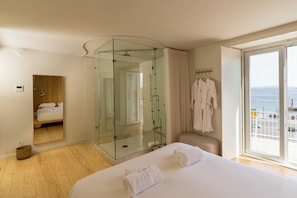 Quarto de hotel com cama, chuveiro envidraçado, vista para o mar e varanda com roupas a secar.