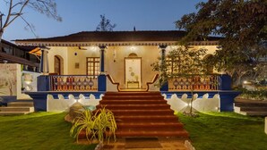 Um edifício com uma escadaria que leva a uma porta, rodeado por um gramado bem cuidado e cerca decorativa.