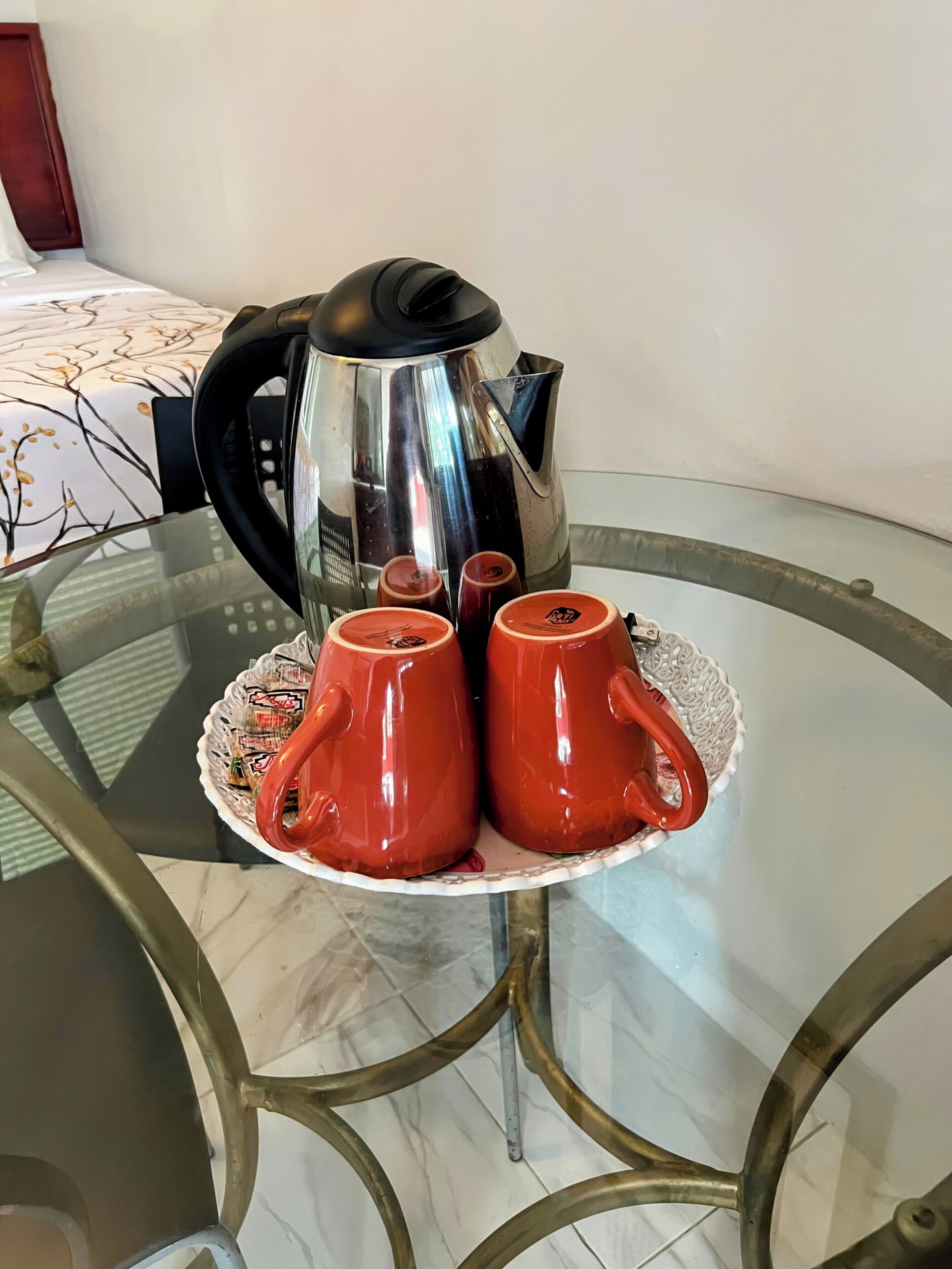 Uma mesa de vidro com estrutura metálica, uma chaleira elétrica de aço inoxidável e duas chávenas de cerâmica vermelhas sobre um tabuleiro branco.