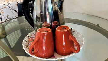 Uma mesa de vidro com estrutura metálica, uma chaleira elétrica de aço inoxidável e duas chávenas de cerâmica vermelhas sobre um tabuleiro branco.
