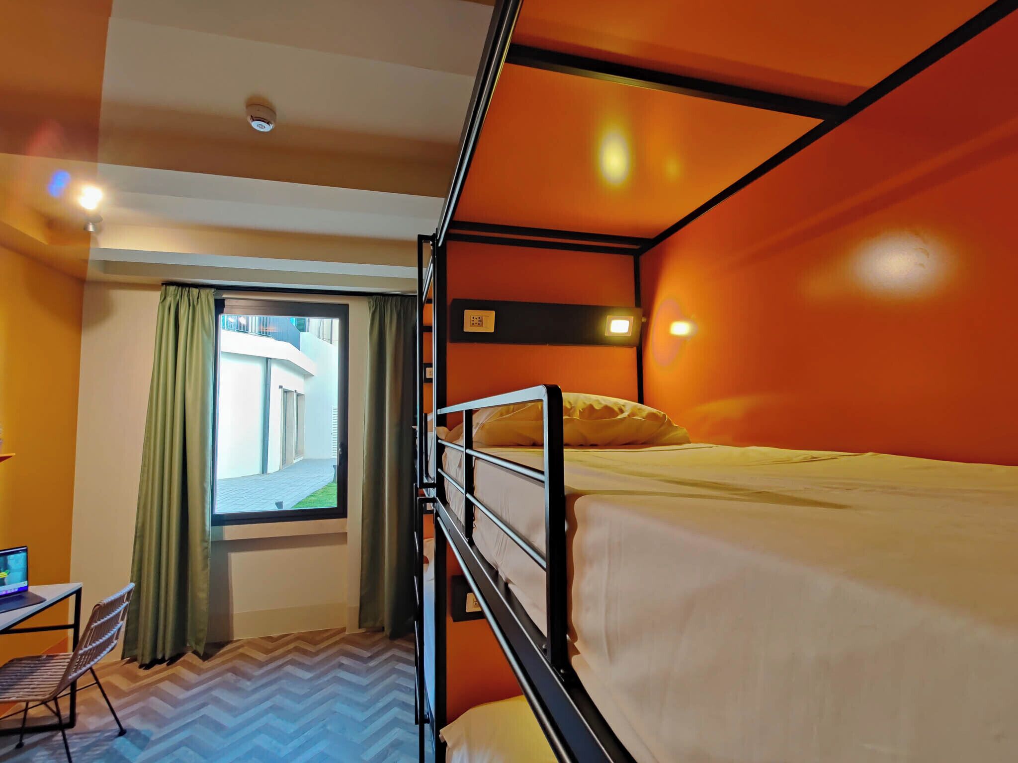 Um quarto de dormitório com beliches, uma escrivaninha com um laptop e uma janela com cortinas.