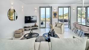 Uma sala de estar moderna com televisão de tela plana, assentos confortáveis, um espaço de jantar com mesa e cadeiras, e janelas amplas com vista para a paisagem.