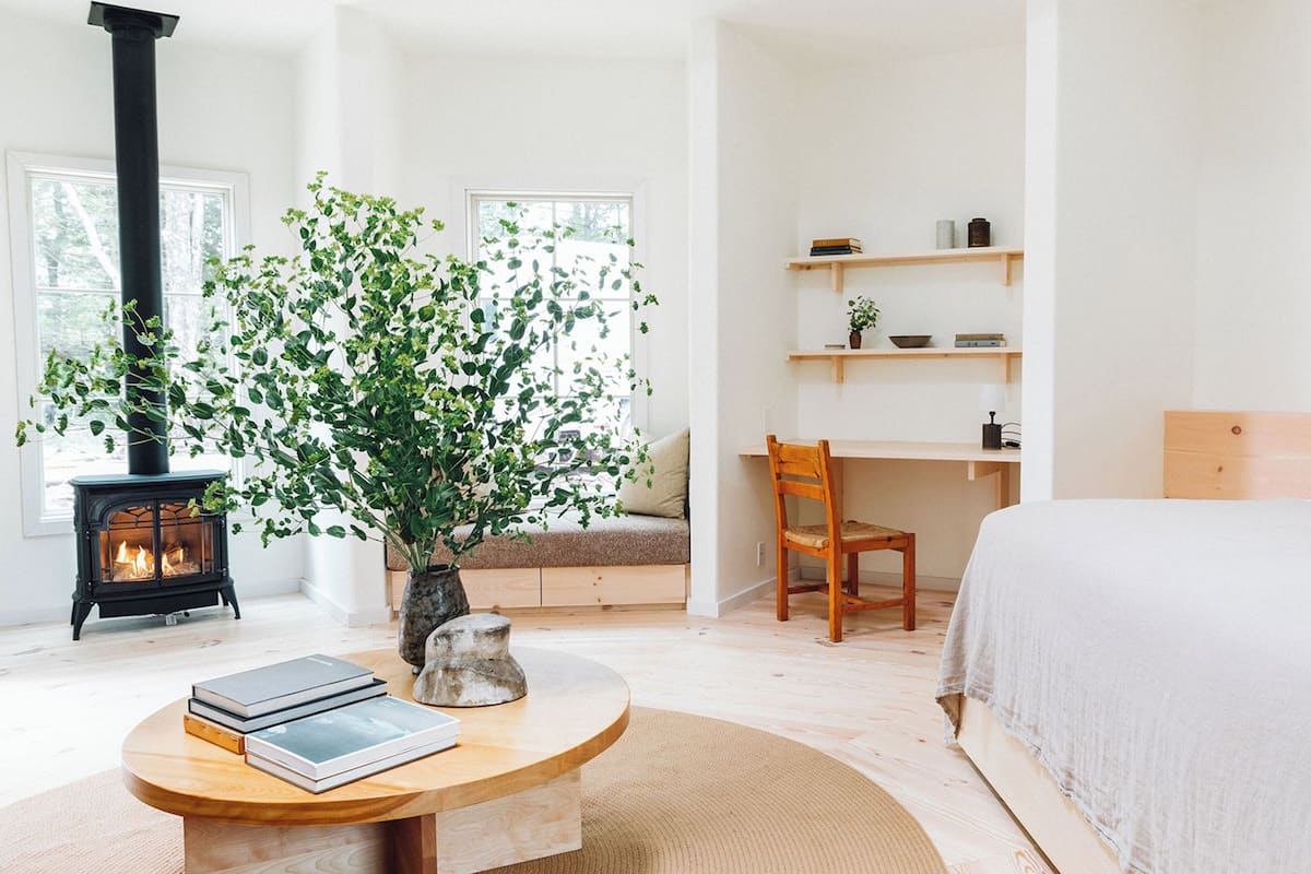 Quarto com cama, escrivaninha, cadeira, mesa redonda com livros, planta em vaso, lareira e estantes com livros e objetos decorativos.