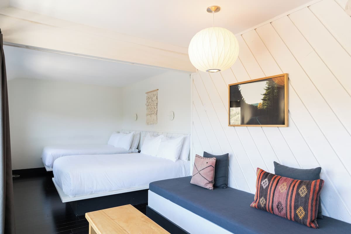 Quarto de hotel com duas camas, um sofá com almofadas, uma mesa de madeira e um candeeiro suspenso.