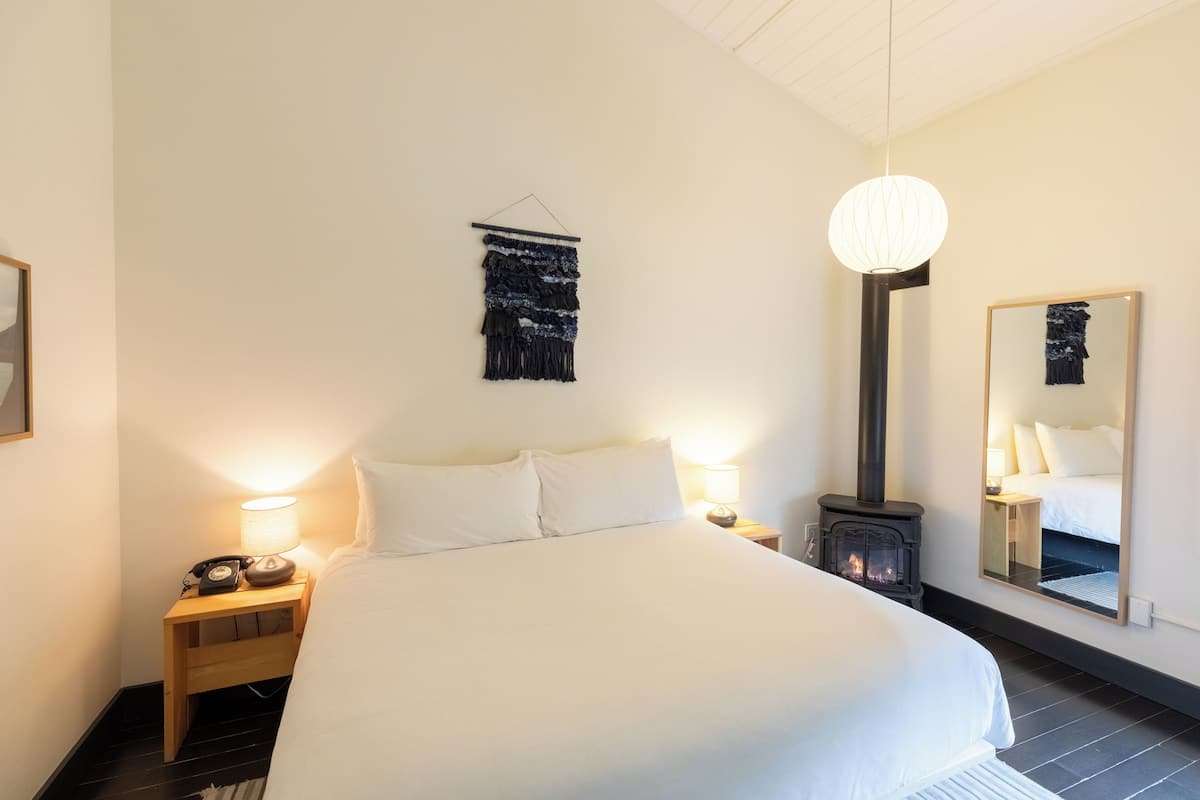 Quarto com uma cama grande, uma mesa de cabeceira de madeira com abajur, tapeçaria na parede, um espelho e uma lareira a lenha.