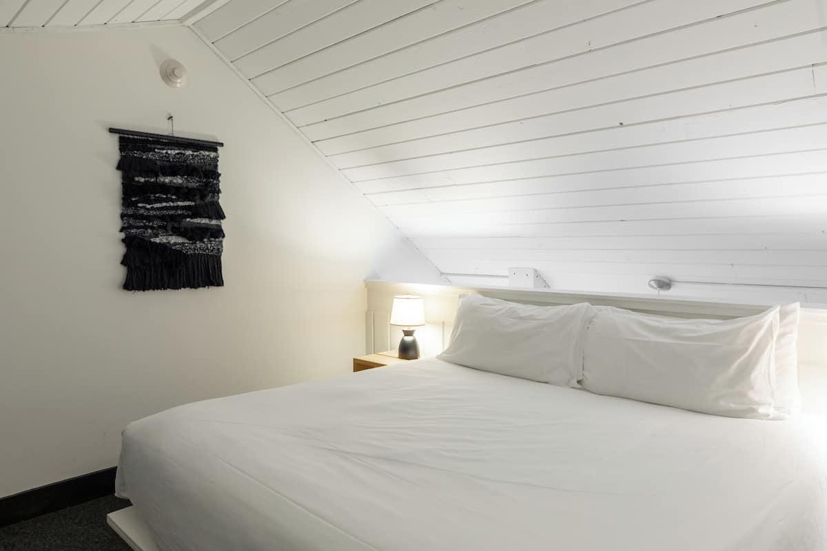 Quarto com cama branca, tapeçaria pendurada, mesa de cabeceira com abajur e teto inclinado.