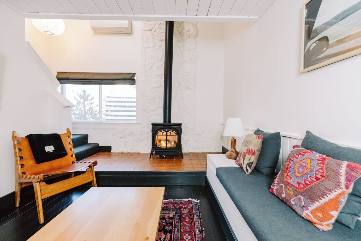 Uma sala de estar aconchegante com lareira, um sofá com almofadas estampadas, uma cadeira de madeira e uma mesa de centro.