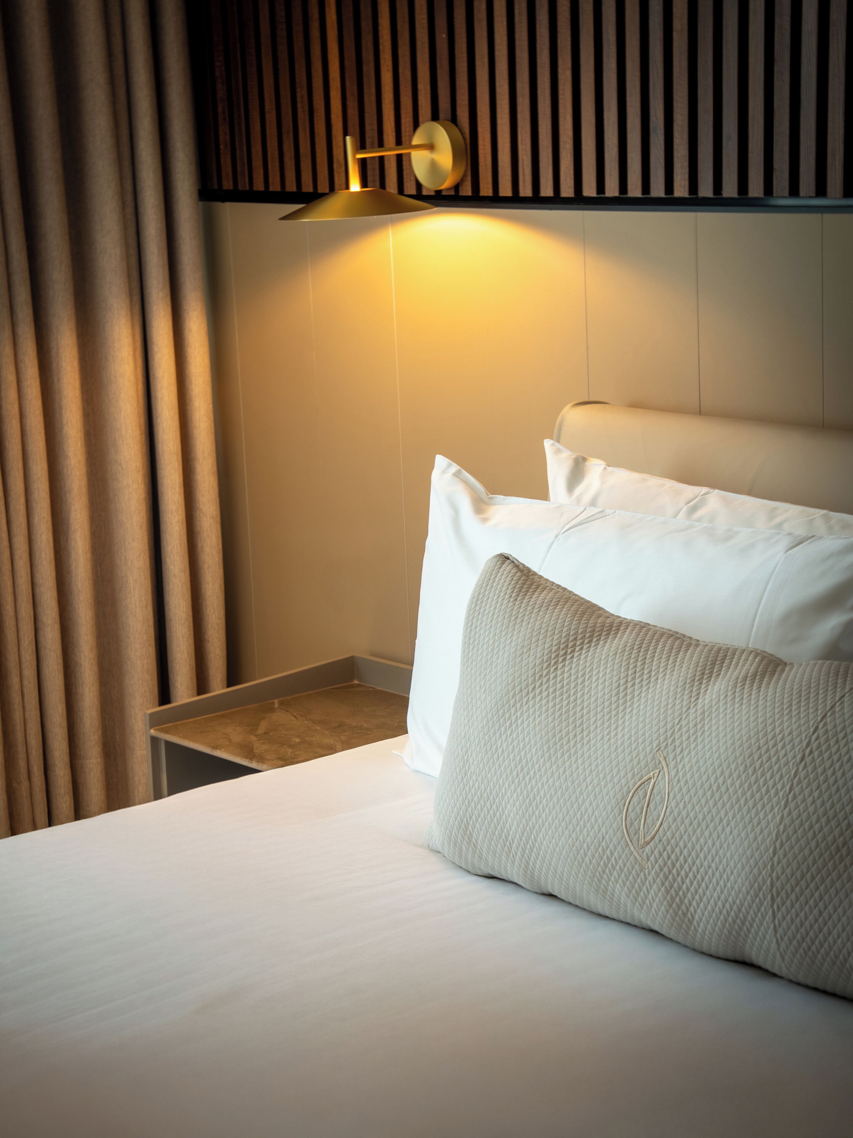 Cama bem arrumada com lençóis brancos e almofada texturizada, mesa de cabeceira com abajur e parede de painel de madeira.