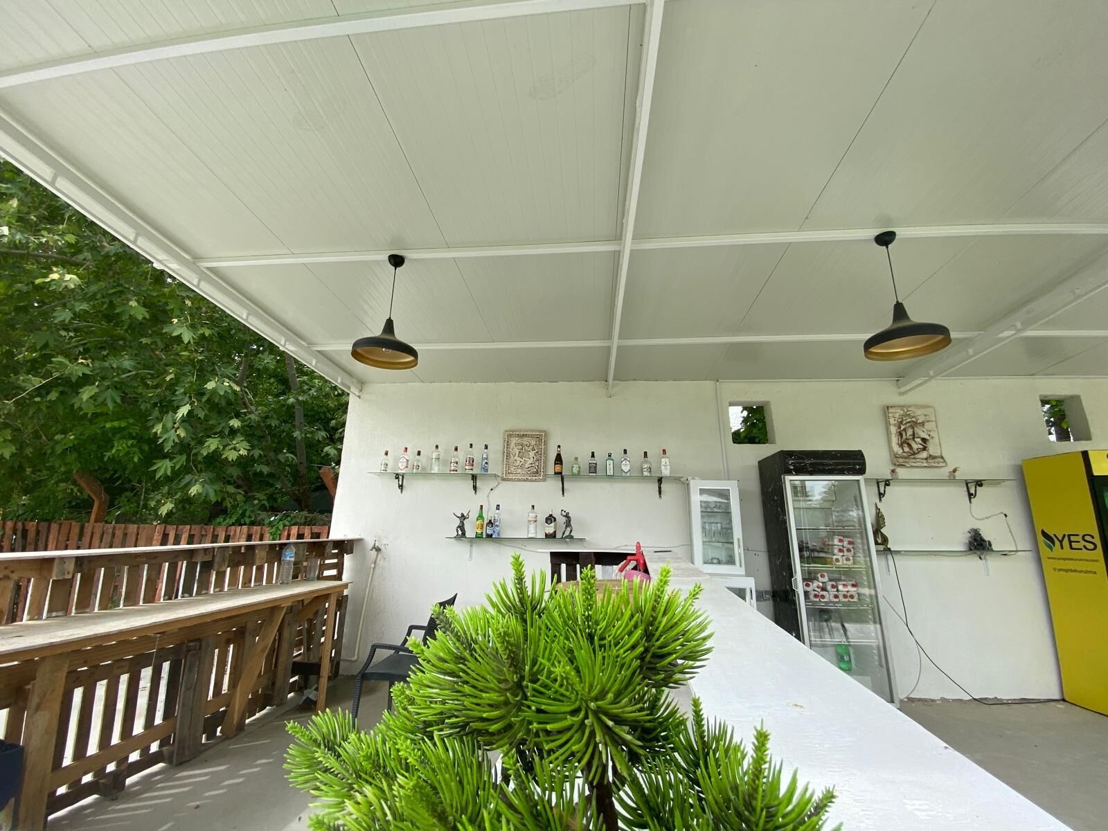Uma área externa coberta com uma mesa comprida, luminárias penduradas e prateleiras com garrafas.