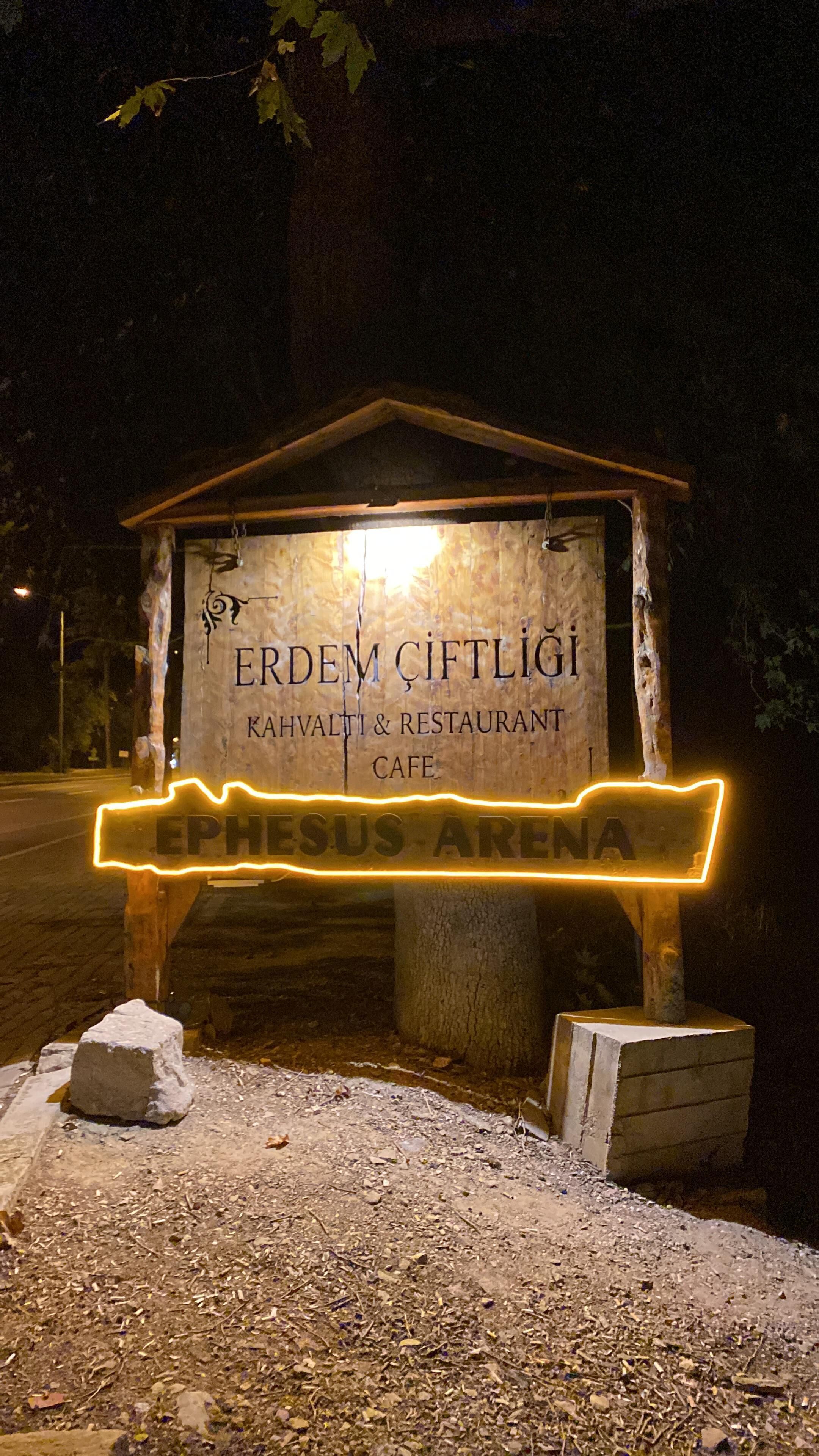 Uma placa de madeira com o texto "ERDEM ÇIFTLİGI KAHVALTI & RESTAURANT CAFE" e "EPHESUS ARENA" iluminada à noite.