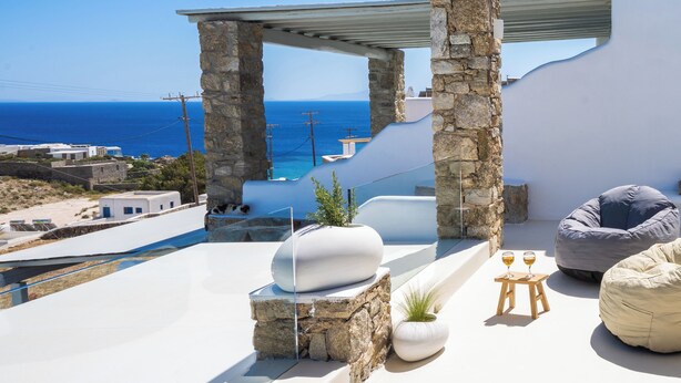 Um terraço moderno ao ar livre com pilares de pedra, uma piscina infinita branca e vista para o mar.